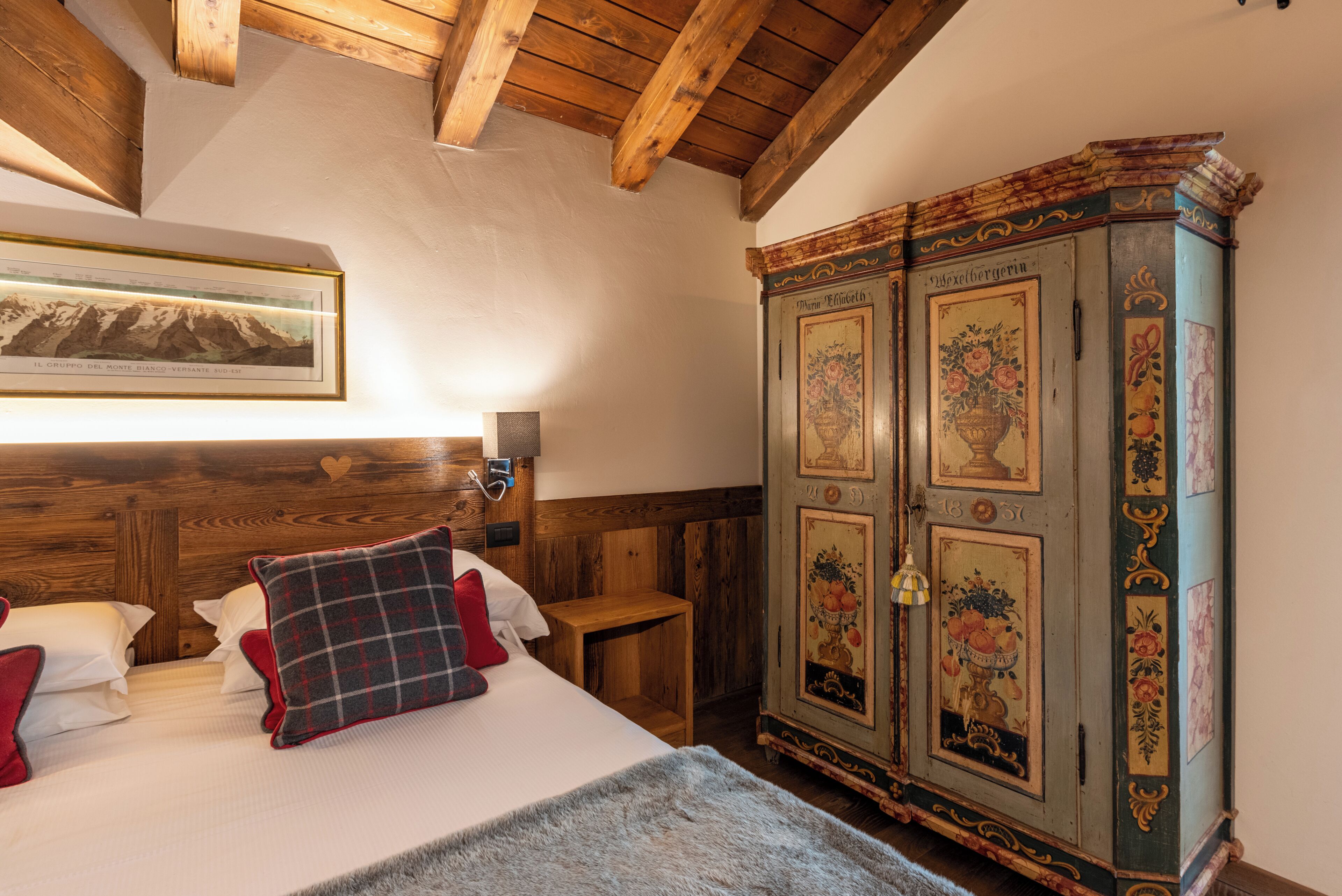 Quarto com cabeceira de madeira, cama com lençóis brancos e almofadas xadrez, mesa de cabeceira de madeira e um aparador antigo com desenhos florais.