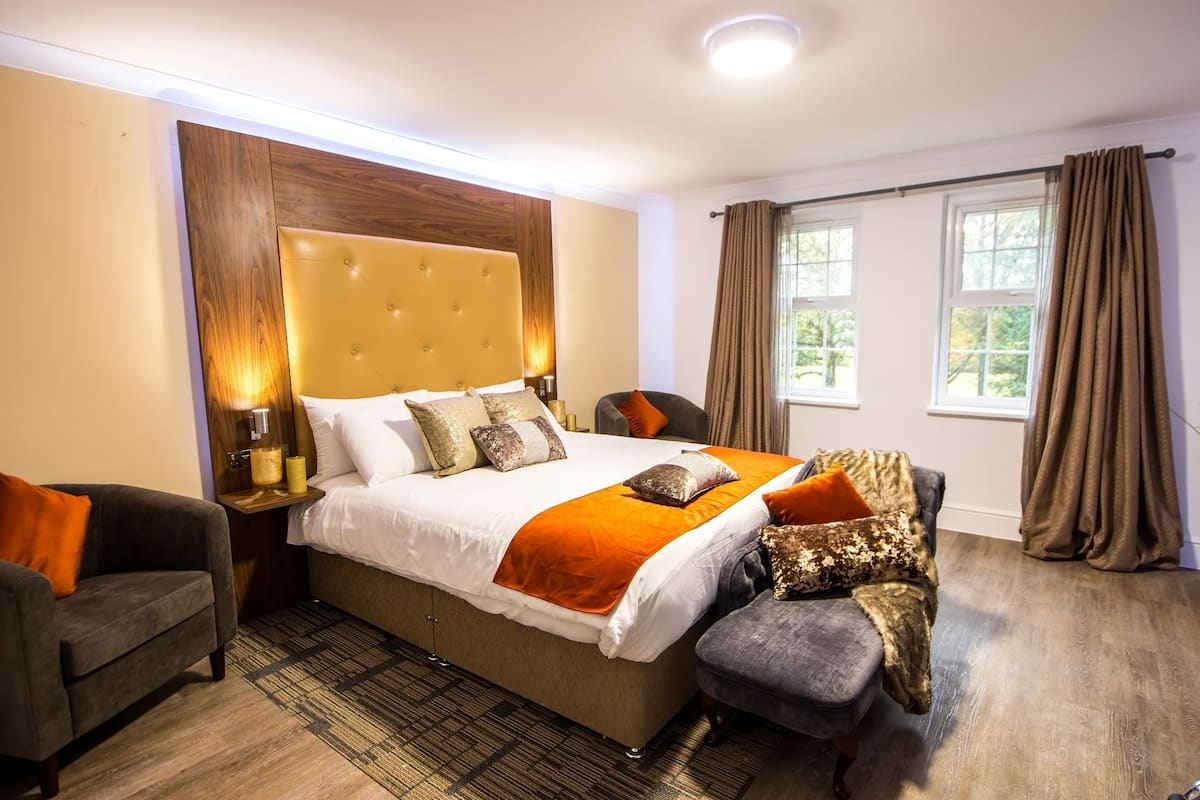 Quarto de hotel com uma cama grande, um sofá, uma mesa de cabeceira e uma janela com cortinas.