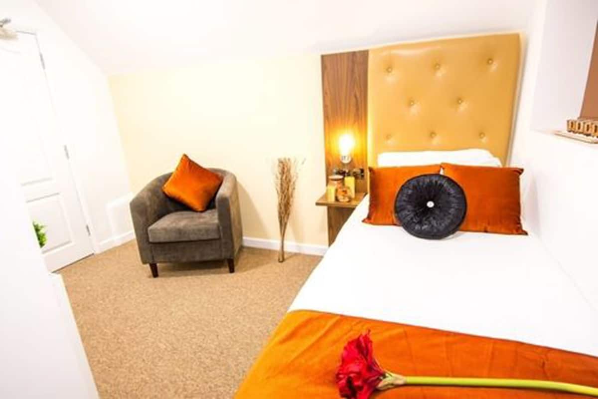 Quarto de hotel com cama, uma cadeira, mesa de cabeceira e luminária fixada na parede.