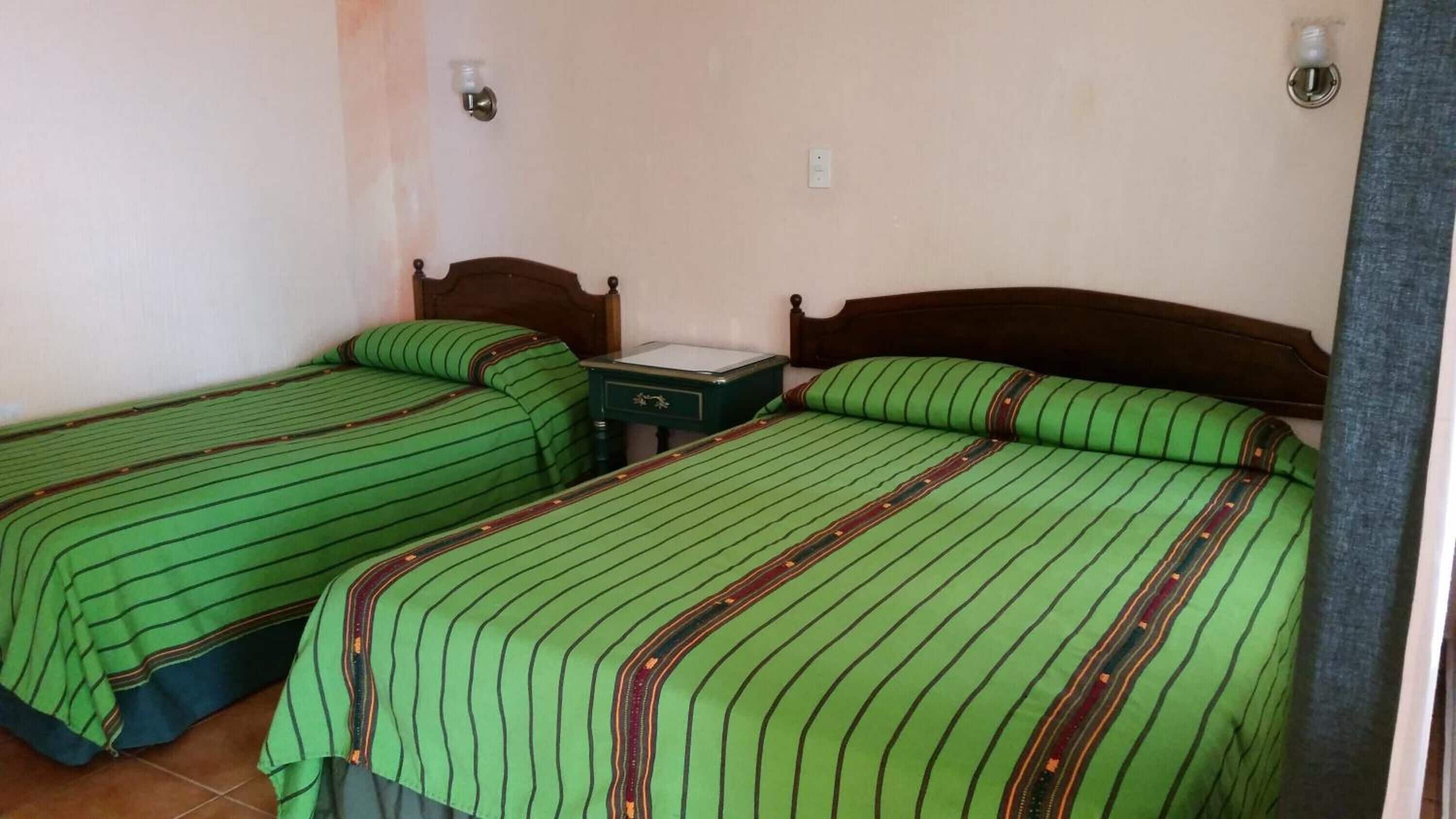 Duas camas de solteiro com roupa de cama verde listrada, cabeceiras de madeira e uma pequena mesa de cabeceira com abajur.