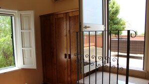 Quarto com cama, guarda-roupa de madeira, varanda com guarda-corpo de metal e janela com vista para o exterior.