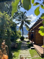 Uma passarela de madeira leva a uma área externa coberta, com uma palmeira e montanhas ao fundo.