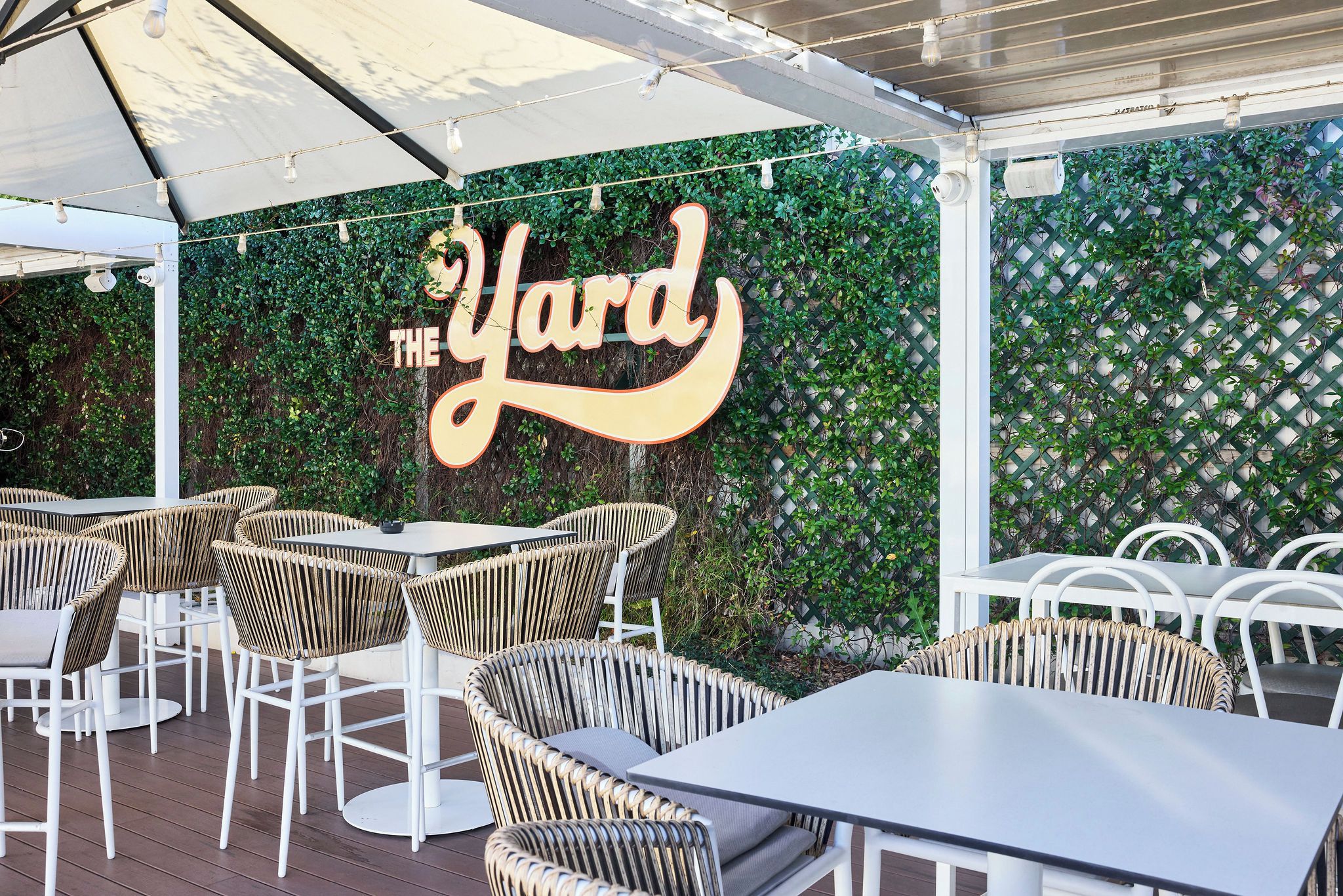 Uma área externa com assentos de vime, mesas, uma placa com o texto 'The Yard' e uma parede verde coberta de folhagem.