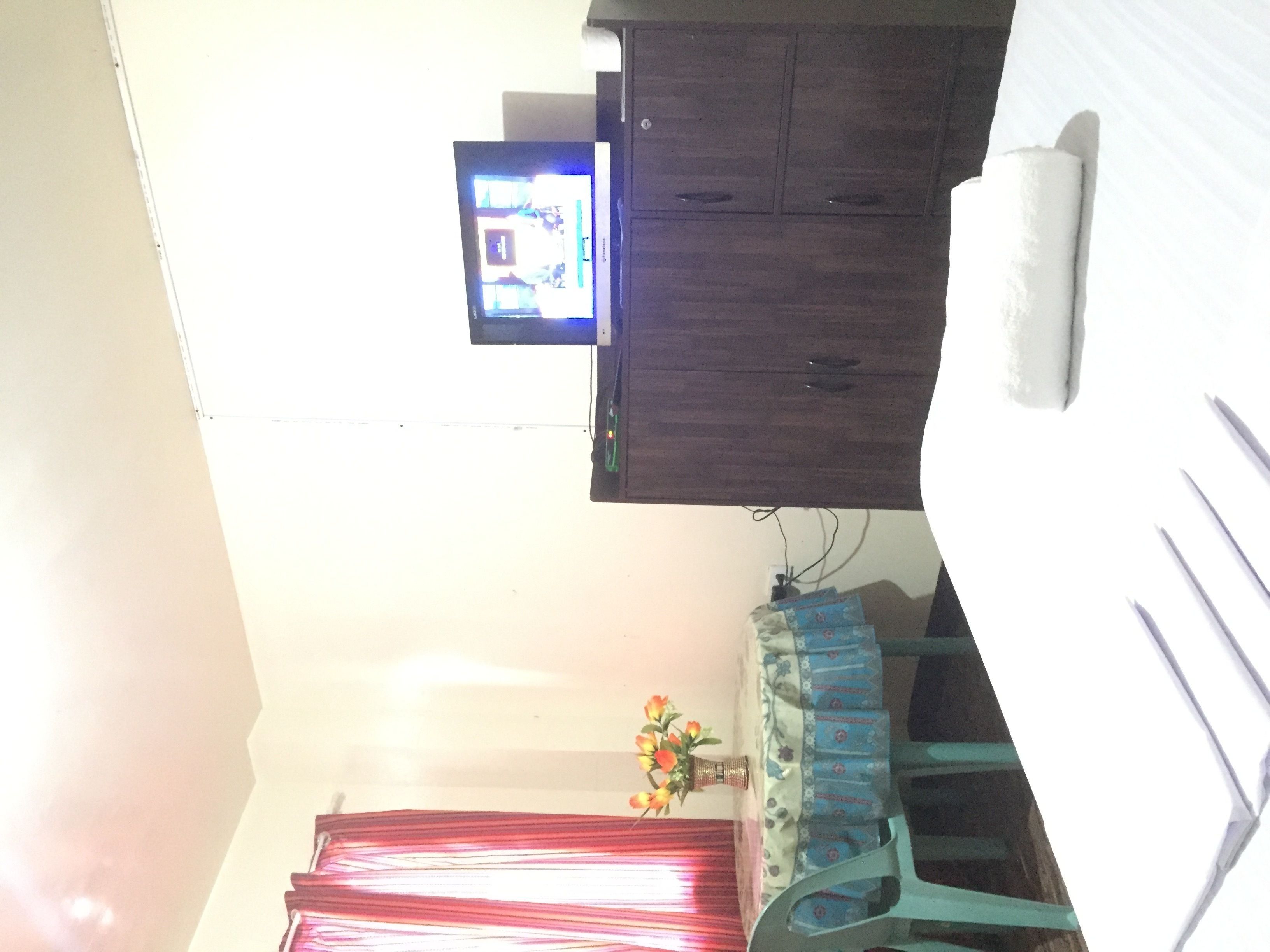 Quarto com televisão fixada na parede, um armário de madeira, parede branca e uma pequena mesa com um vaso de flores.