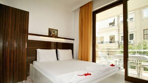 Quarto de hotel com uma cama grande, cabeceira de madeira, varanda com mesa e cadeiras, e vista para o edifício.