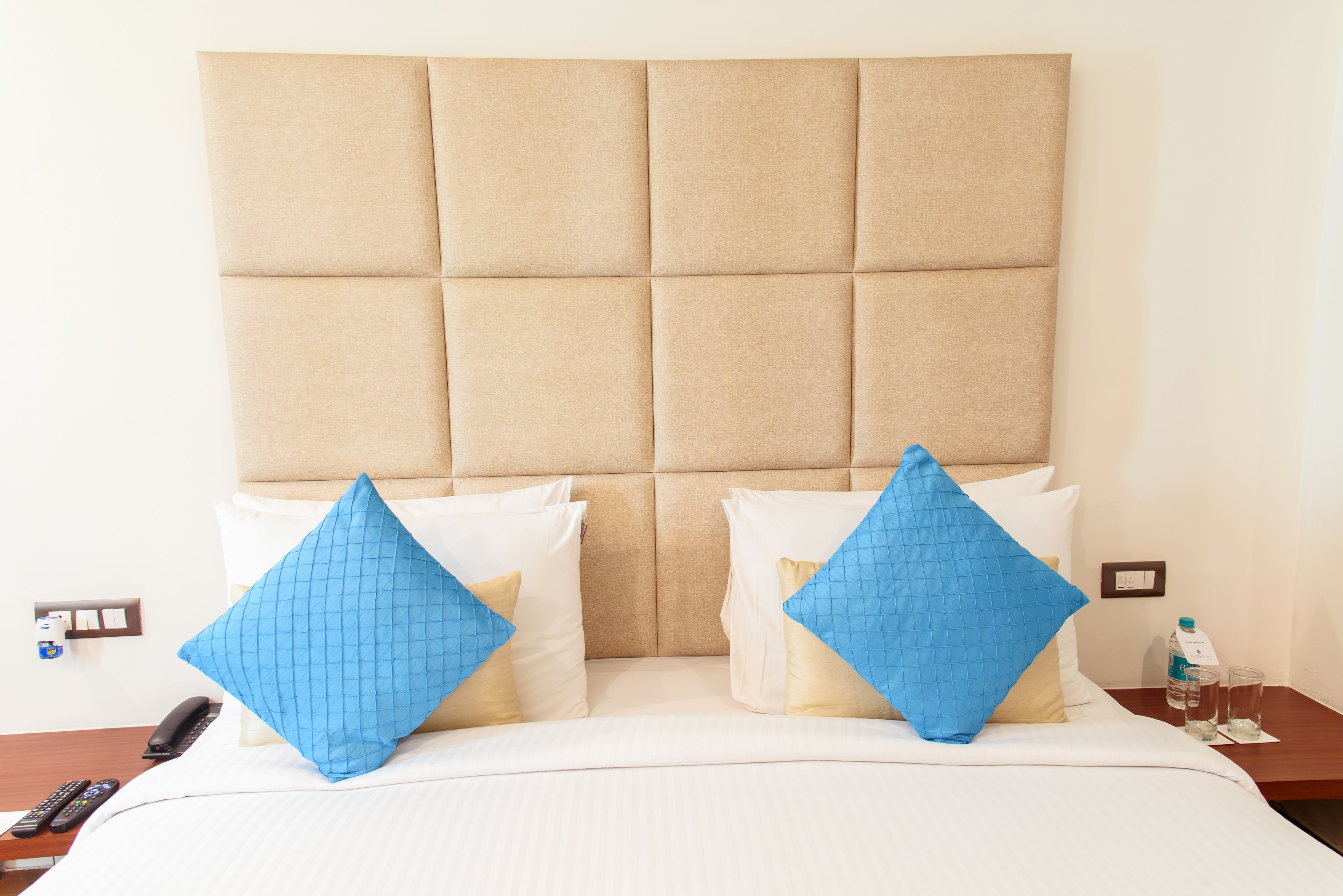 Quarto de hotel com cabeceira estofada, duas almofadas azuis, espalha branca na cama, mesa de cabeceira de madeira com um controle remoto, uma garrafa de água e um copo.
