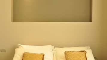 Cama bem arrumada com duas almofadas, bandeja de café da manhã com croissants, frutas e suco de laranja, e luminária fixada na parede.