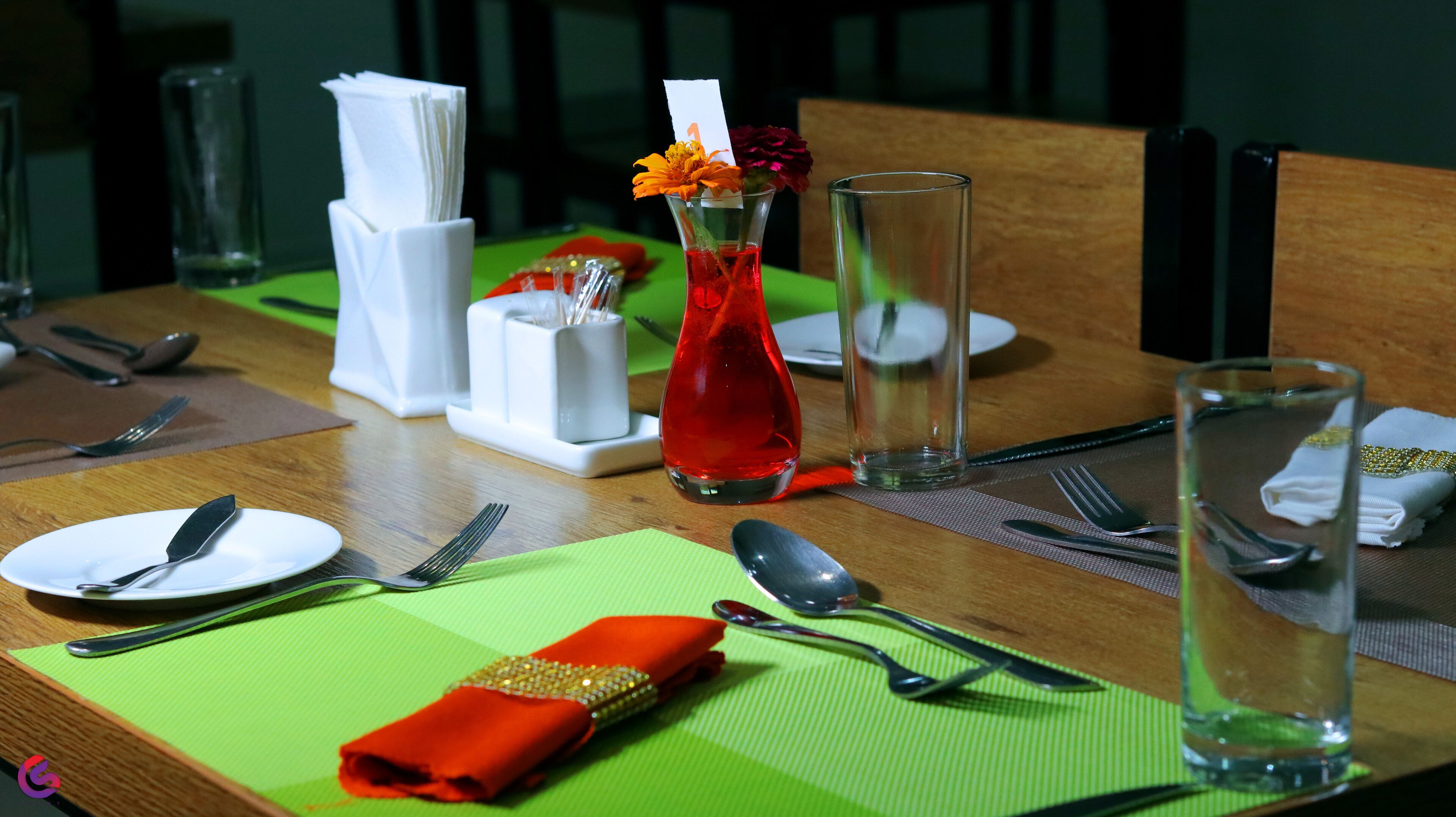 Mesa posta com pratos, talheres, um copo de água, um vaso vermelho com flores e um porta-guardanapos.