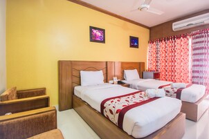 Quarto de hotel com duas camas, cabeceiras de madeira, ventoinha no teto, ar condicionado e janela com cortinas estampadas.