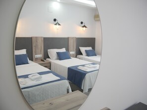Vista refletida de um quarto de hotel com três camas, cada uma com colcha azul e branca, cabeceira de madeira e mesa de cabeceira com telefone e controle remoto.