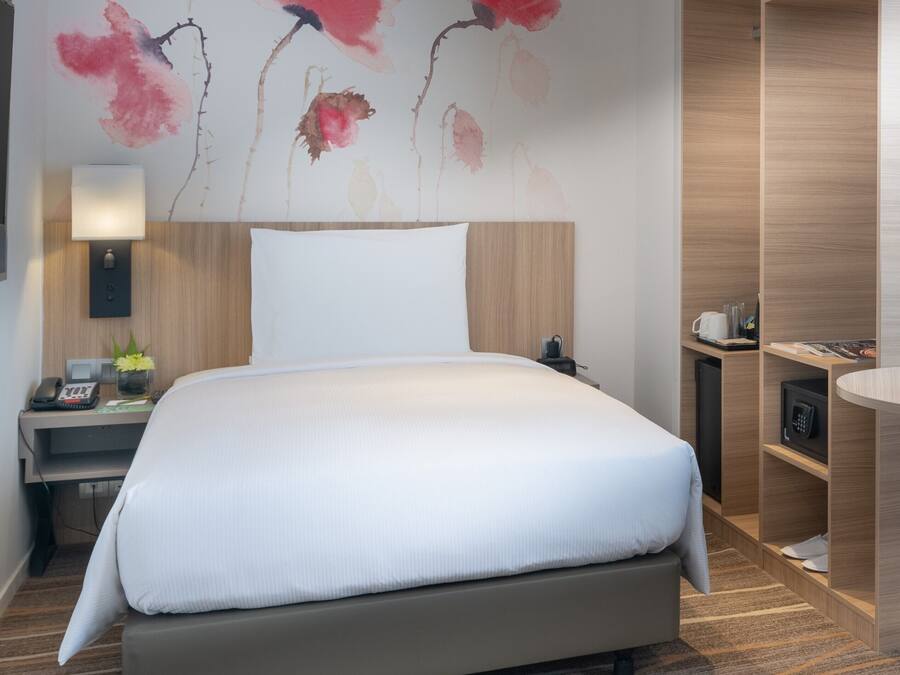 Quarto de hotel com uma cama grande, uma mesa de cabeceira com abajur, telefone e uma pequena planta. Há um guarda-roupa de madeira e uma escrivaninha com cadeira.