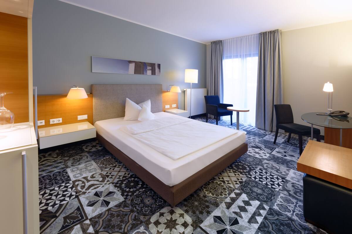 Quarto de hotel com uma cama grande, duas mesas de cabeceira com luminárias, uma escrivaninha com cadeira e um tapete estampado.
