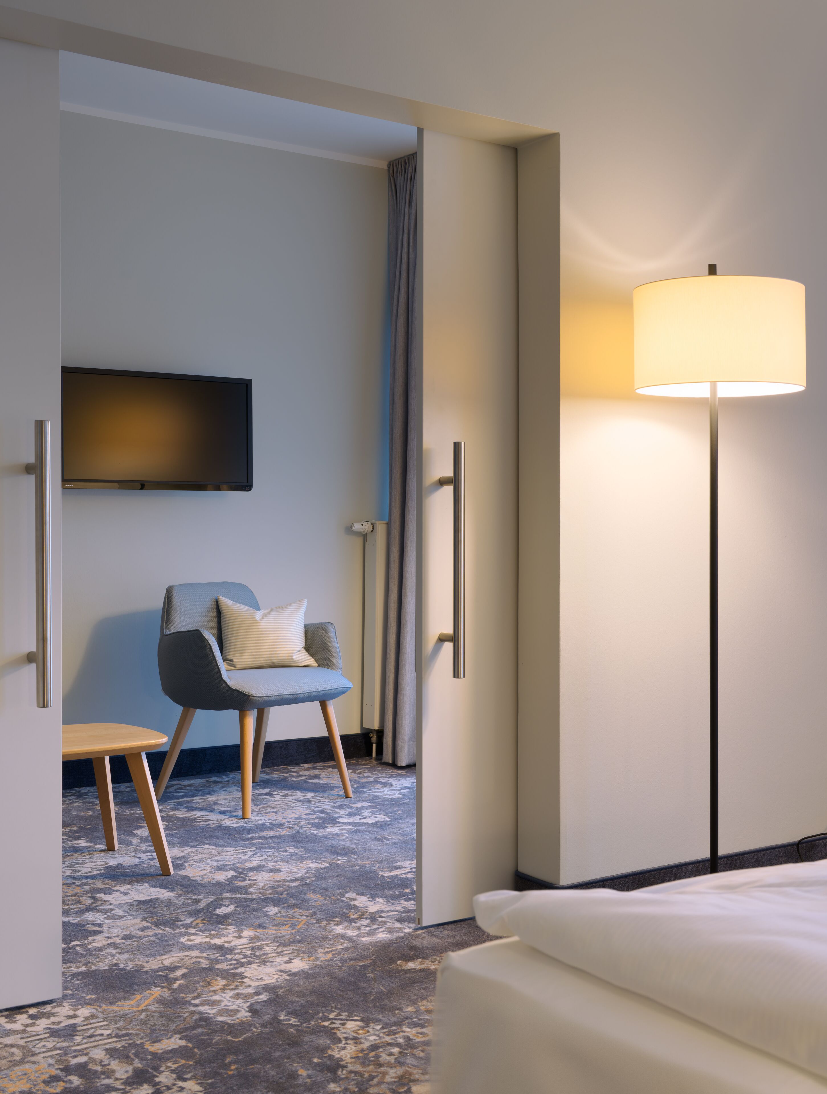 Quarto de hotel com uma cama, uma cadeira com travesseiro, uma TV de tela plana na parede e um abajur.