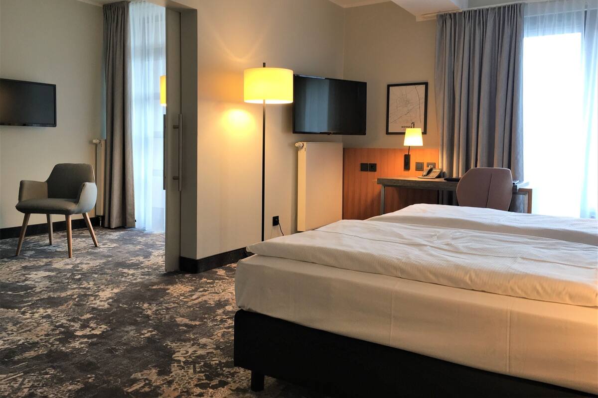 Quarto de hotel com uma cama grande, uma escrivaninha com cadeira, uma televisão na parede e uma janela com cortinas.