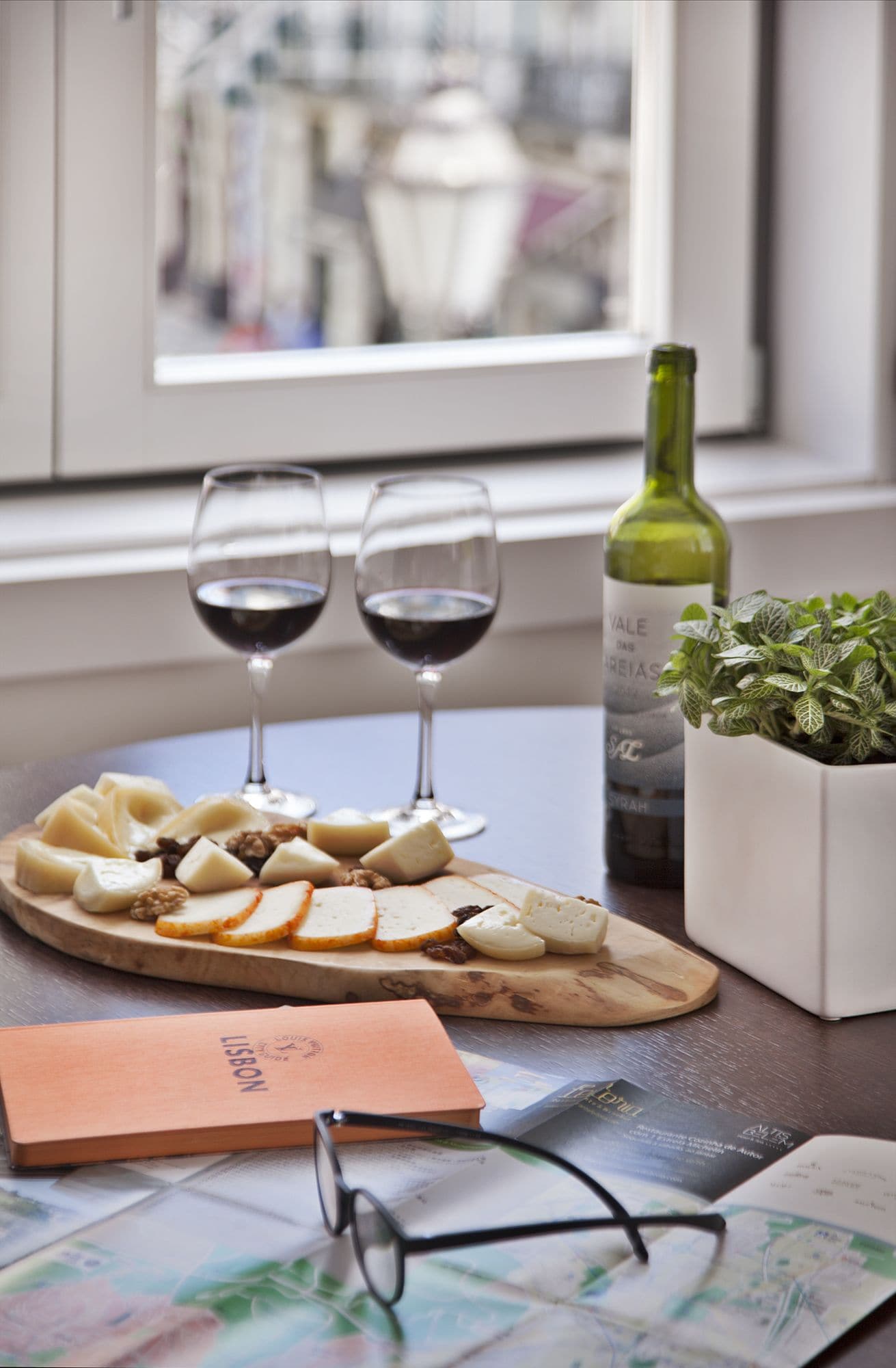 Uma mesa com uma tábua de queijos, dois copos de vinho tinto, uma garrafa de vinho, uma planta em vaso, um livro e um par de óculos sobre um mapa.