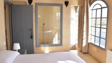 Quarto de hotel com uma cama grande, banheiro com chuveiro, janela com cortinas e vista para o mar.