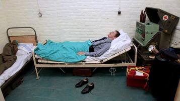 Quarto individual com cama de estrutura metálica, manta azul-esverdeada, um par de sapatos, uma caixa de ferramentas vermelha e um aparelho médico sobre uma mesa.