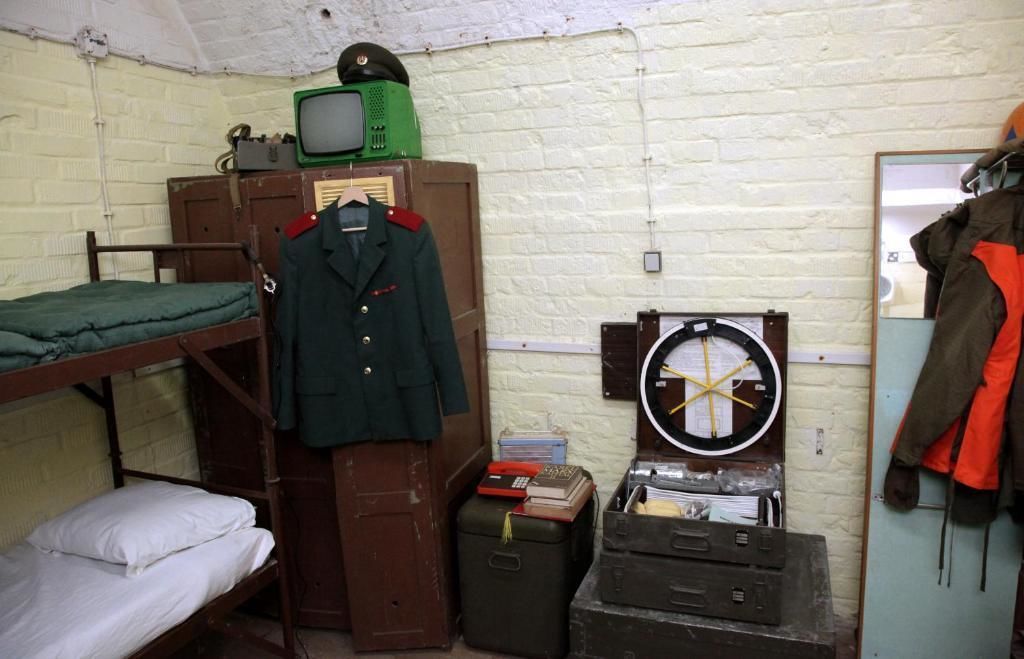 Quarto com beliche, um uniforme verde pendurado na parede, uma televisão pequena e uma mala.