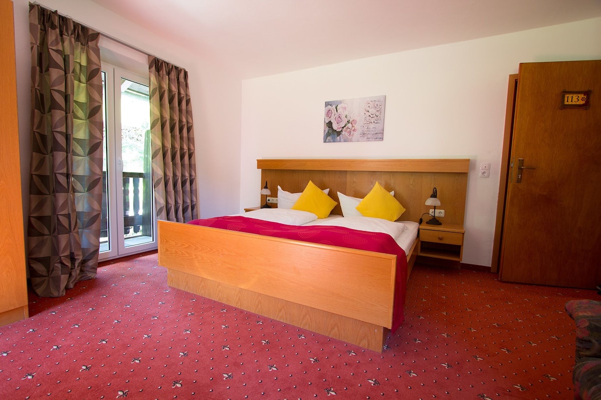 Quarto de hotel com cama, mesa de cabeceira, abajur, porta e janela com cortinas.