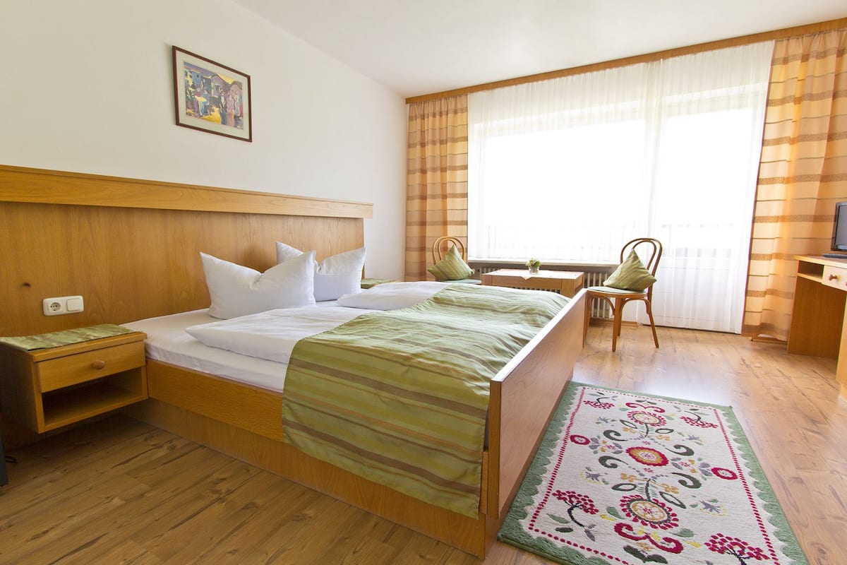 Quarto de hotel com cama de madeira, mesa de cabeceira, escrivaninha, cadeira e uma janela grande com cortinas.