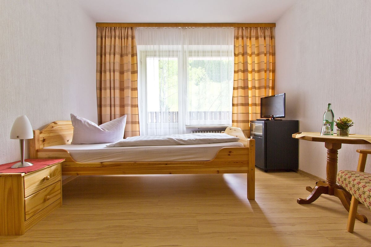 Quarto com cama de madeira, escrivaninha com televisão, cadeira e pequena mesa com garrafa e flores.