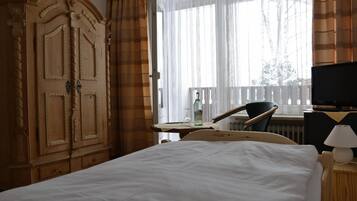 Quarto de hotel com um grande guarda-roupa de madeira, uma cama com roupa de cama branca, uma pequena mesa com uma garrafa, uma cadeira e uma televisão sobre um suporte.