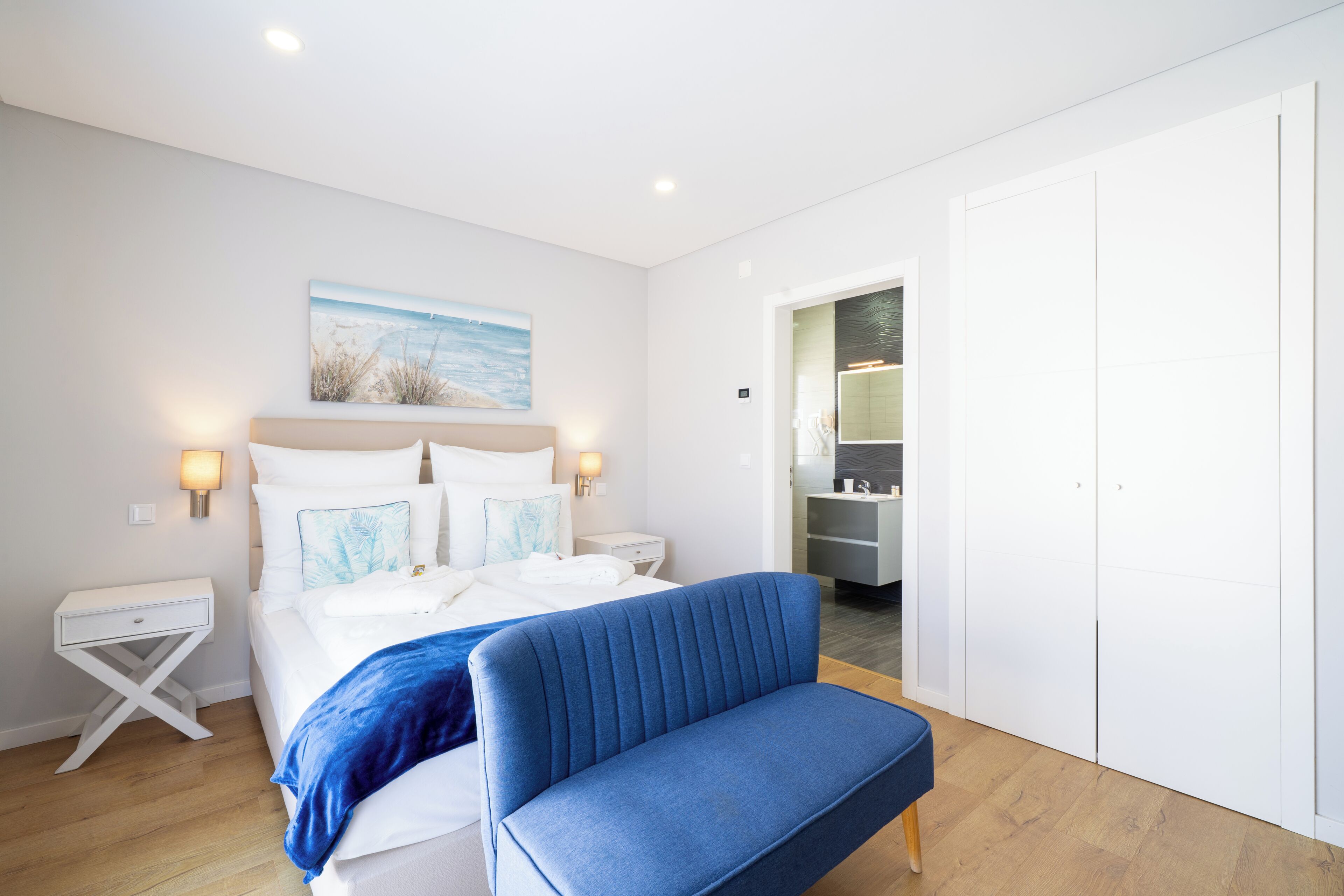 Quarto com cama, mesas de cabeceira, um banco azul, um quadro e um guarda-roupa.