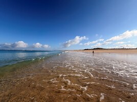 Praia com água cristalina, uma pessoa caminhando e edifícios ao longe.