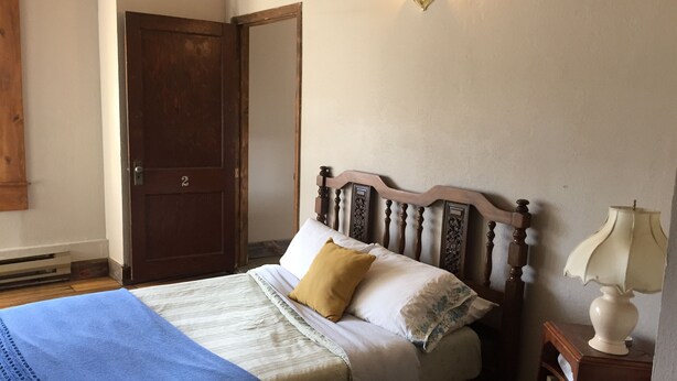 Quarto com cama de madeira, cobertor azul, travesseiro branco e travesseiro amarelo, luminária fixada na parede e porta com número.