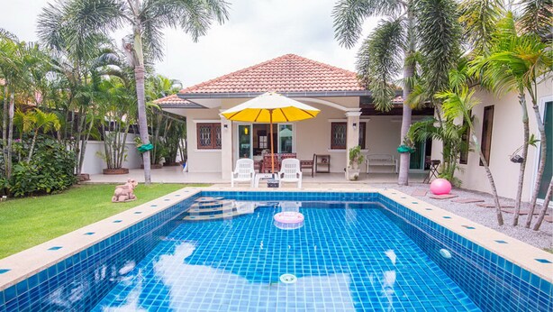 Uma piscina com deck revestido, área de estar coberta com cadeiras e um guarda-sol, e uma casa com telhado telhado ao fundo.