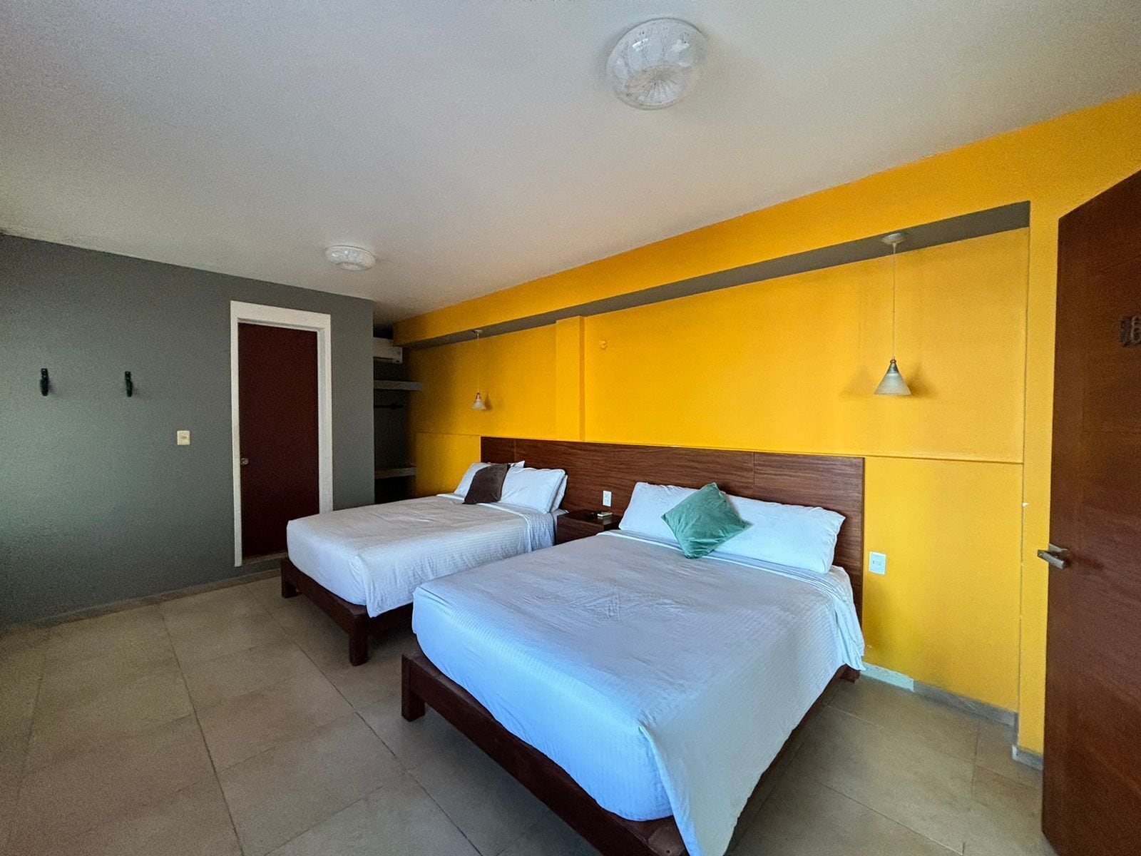Quarto de hotel com duas camas, cabeceira de madeira, uma porta e uma parede amarela como detalhe.