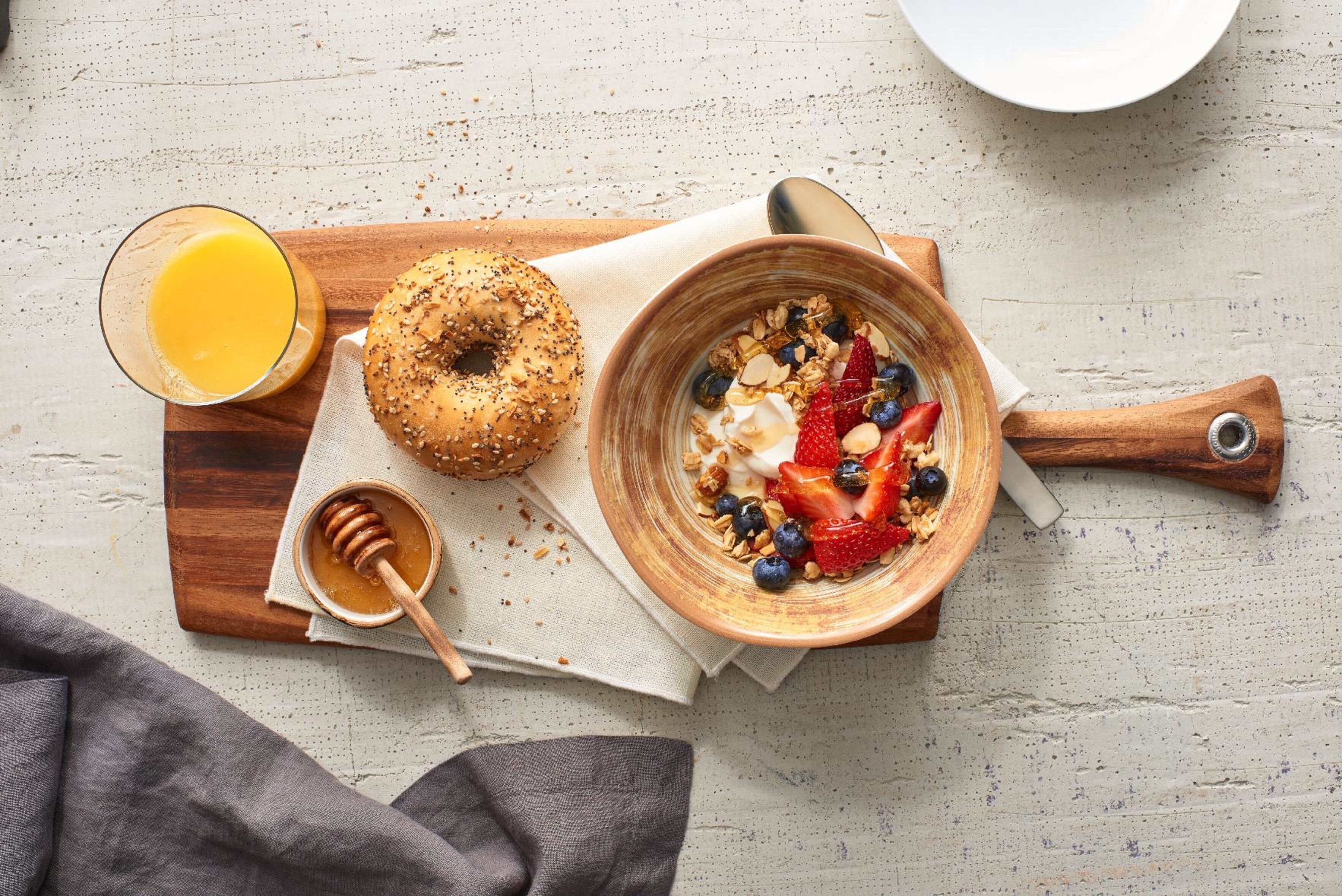 Uma mesa de pequeno-almoço com iogurte, frutas, granola, sumo de laranja, um bagel e um pires de mel sobre uma tábua de madeira.