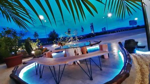 Uma área externa com mesas e cadeiras, cercada por plantas em vasos e uma piscina decorativa.