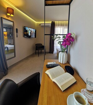 Quarto de hotel com cama, escrivaninha, um livro, controle remoto, um copo d'água e um vaso com flores.