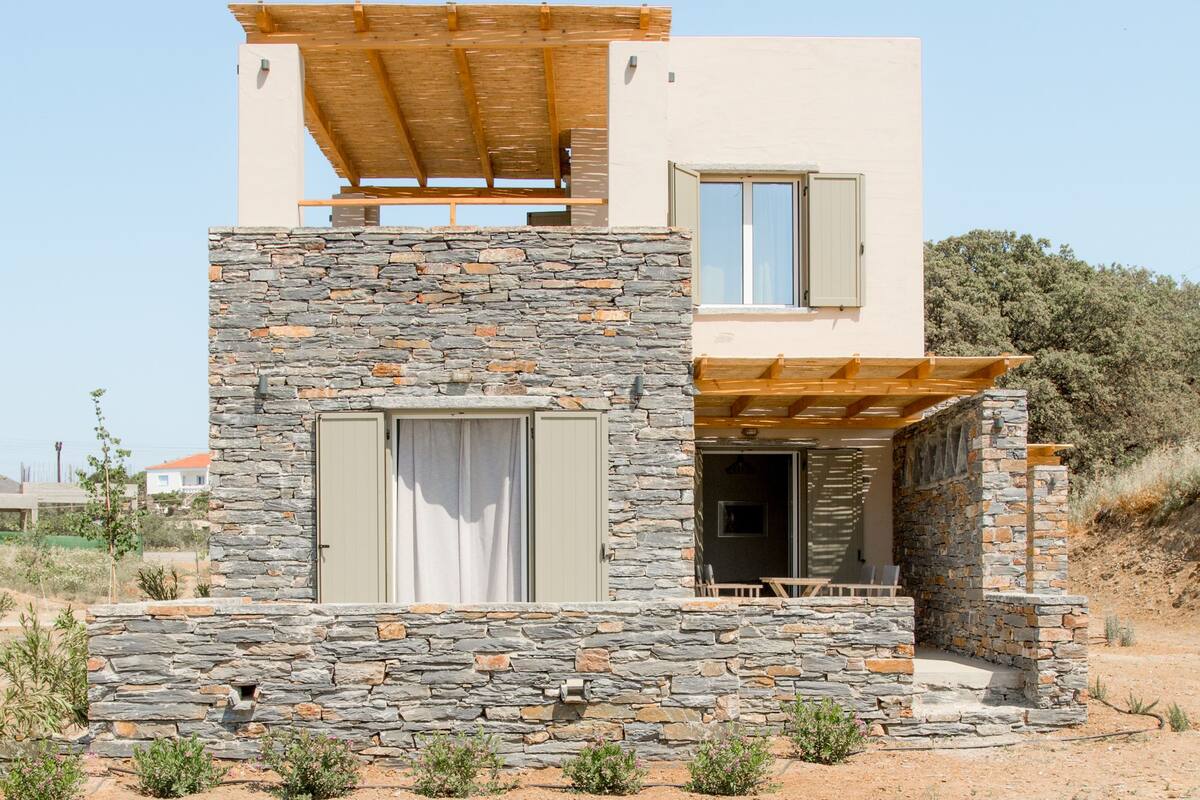 Uma casa moderna de dois andares com fachada de pedra, uma pérgula de madeira no andar superior e uma pequena varanda.