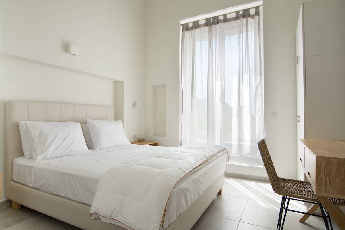 Quarto com cama, uma cadeira, mesa de cabeceira e janela com cortinas leves.