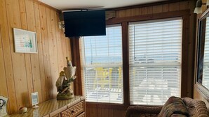 Quarto com paredes de madeira, televisão presa à parede, sofá de vime e janela com persianas.