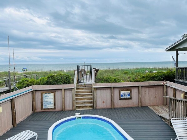 Um deck de madeira com piscina, uma escadaria para uma praia e uma placa de salva-vidas.