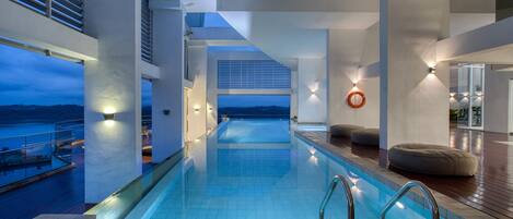Piscina interior moderna com água azul cristalina, rodeada por paredes brancas e grandes janelas.