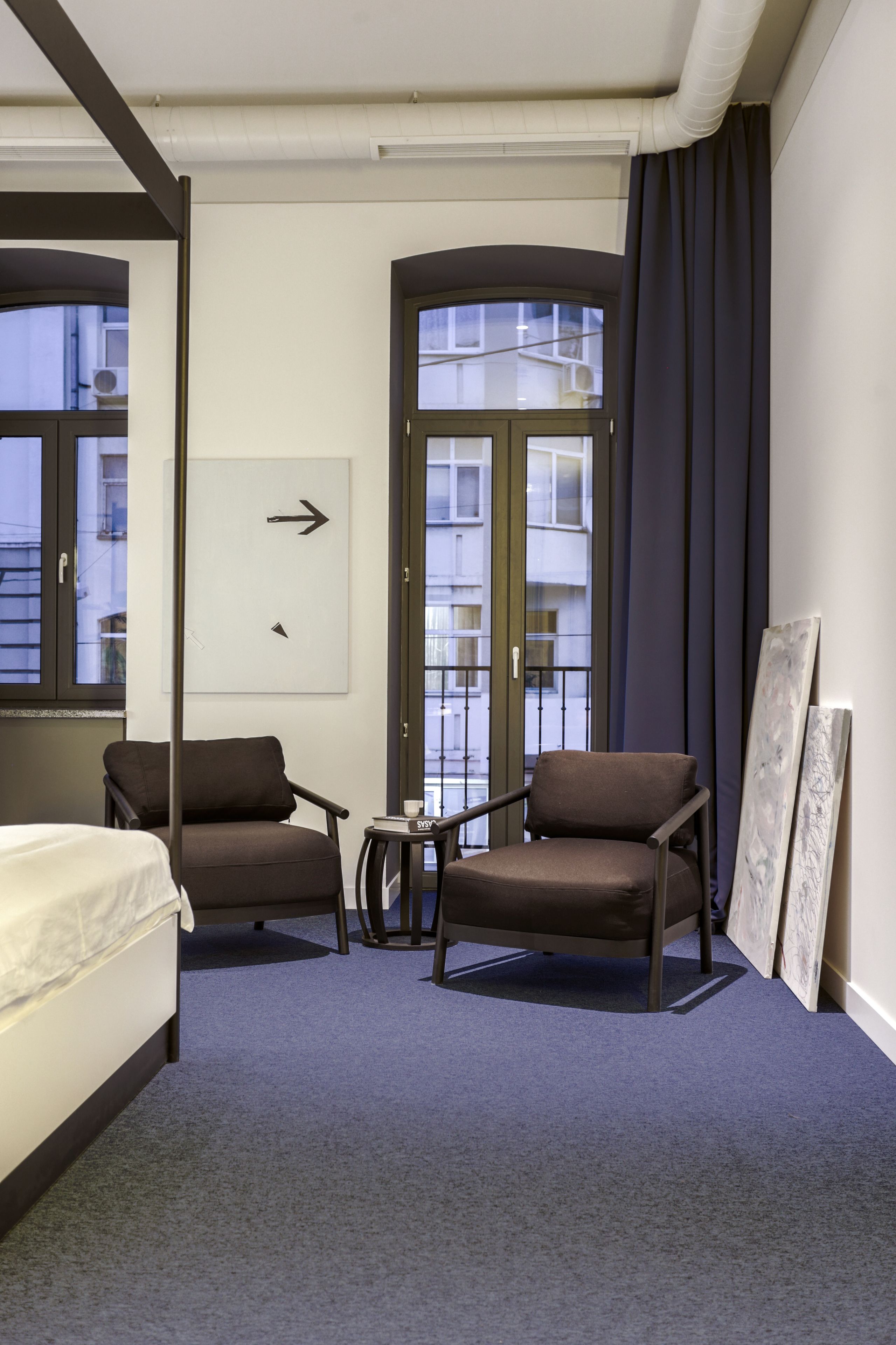 Quarto de hotel com uma cama, duas poltronas, uma mesinha e uma janela grande com cortinas.