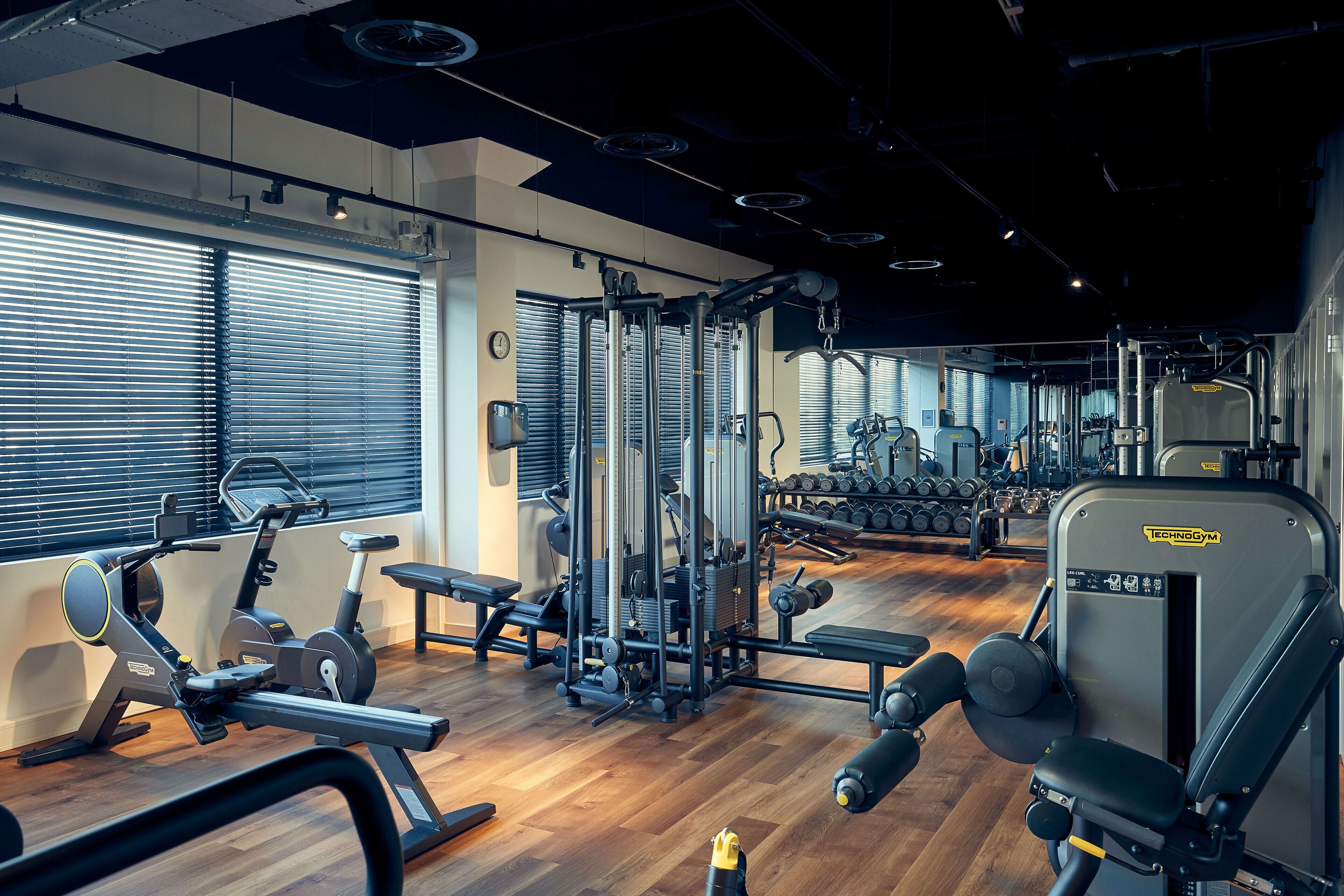 Uma academia com diversos equipamentos de exercício, piso de madeira e grandes janelas com persianas.