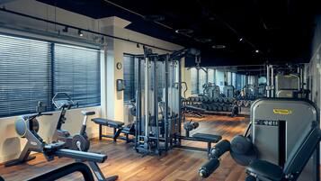 Uma academia com diversos equipamentos de exercício, piso de madeira e grandes janelas com persianas.