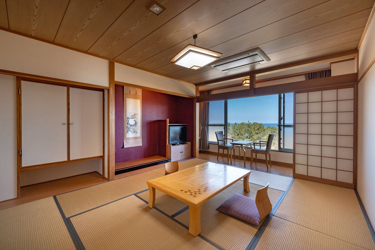 Um quarto japonês tradicional com piso de tatame, uma mesa de madeira baixa, uma televisão e uma porta deslizante que dá para uma varanda com vista.