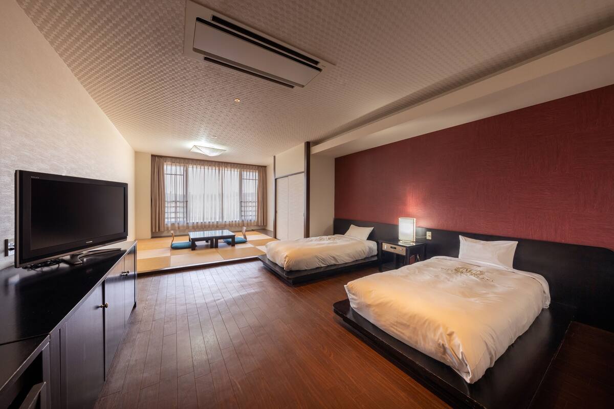 Quarto de hotel com duas camas, uma televisão e uma janela grande com cortinas.