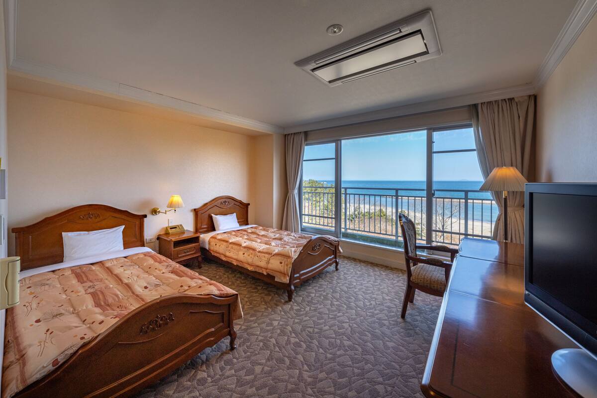 Quarto de hotel com duas camas, televisão, escrivaninha e uma grande janela com vista para o mar.