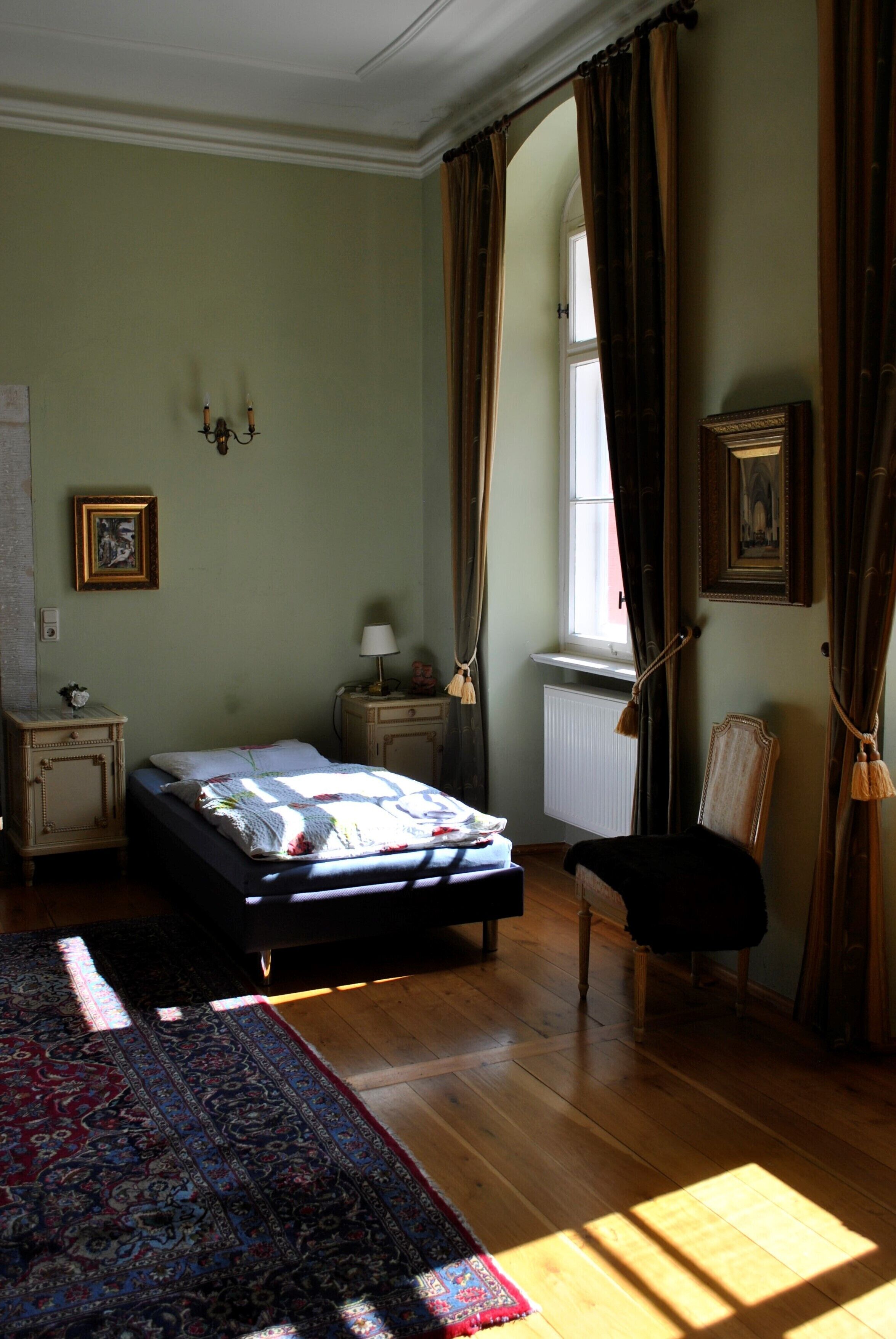 Quarto com cama, cadeira, mesa de cabeceira, luminária, janela com cortinas, radiador e tapete.