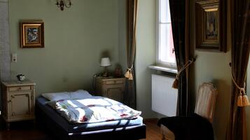 Quarto com cama, cadeira, mesa de cabeceira, luminária, janela com cortinas, radiador e tapete.