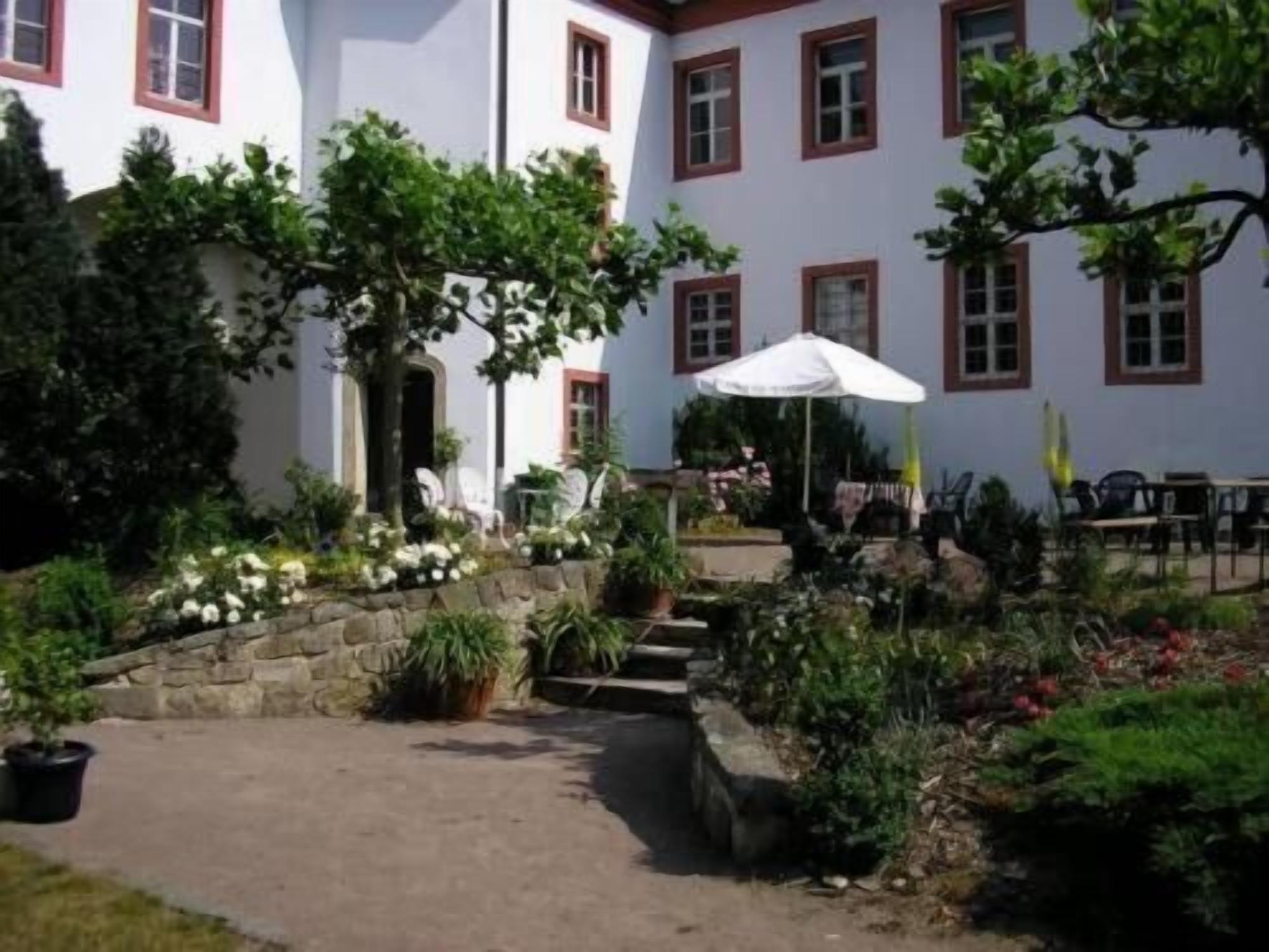 Um edifício com fachada branca e molduras de janelas vermelhas, um pátio pavimentado com plantas em vasos e mesas ao ar livre.