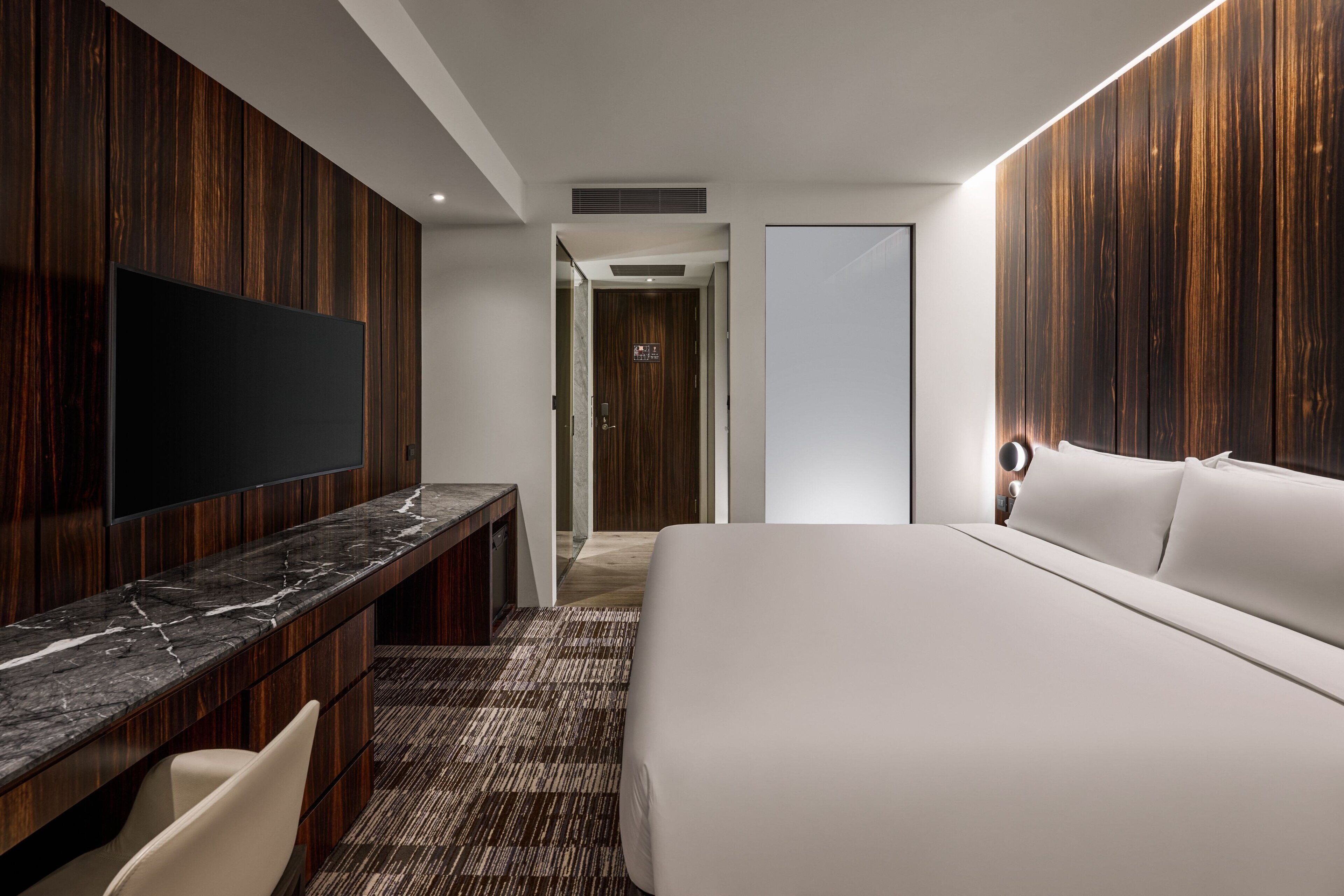 Quarto de hotel moderno com uma cama grande, uma TV de tela plana montada em uma parede revestida de madeira, uma escrivaninha com cadeira e piso coberto de carpete.