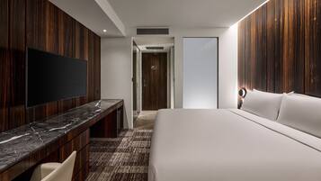 Quarto de hotel moderno com uma cama grande, uma TV de tela plana montada em uma parede revestida de madeira, uma escrivaninha com cadeira e piso coberto de carpete.